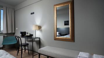 Quarto de hotel com cama, escrivaninha com abajur, cadeira, banco e um espelho grande.