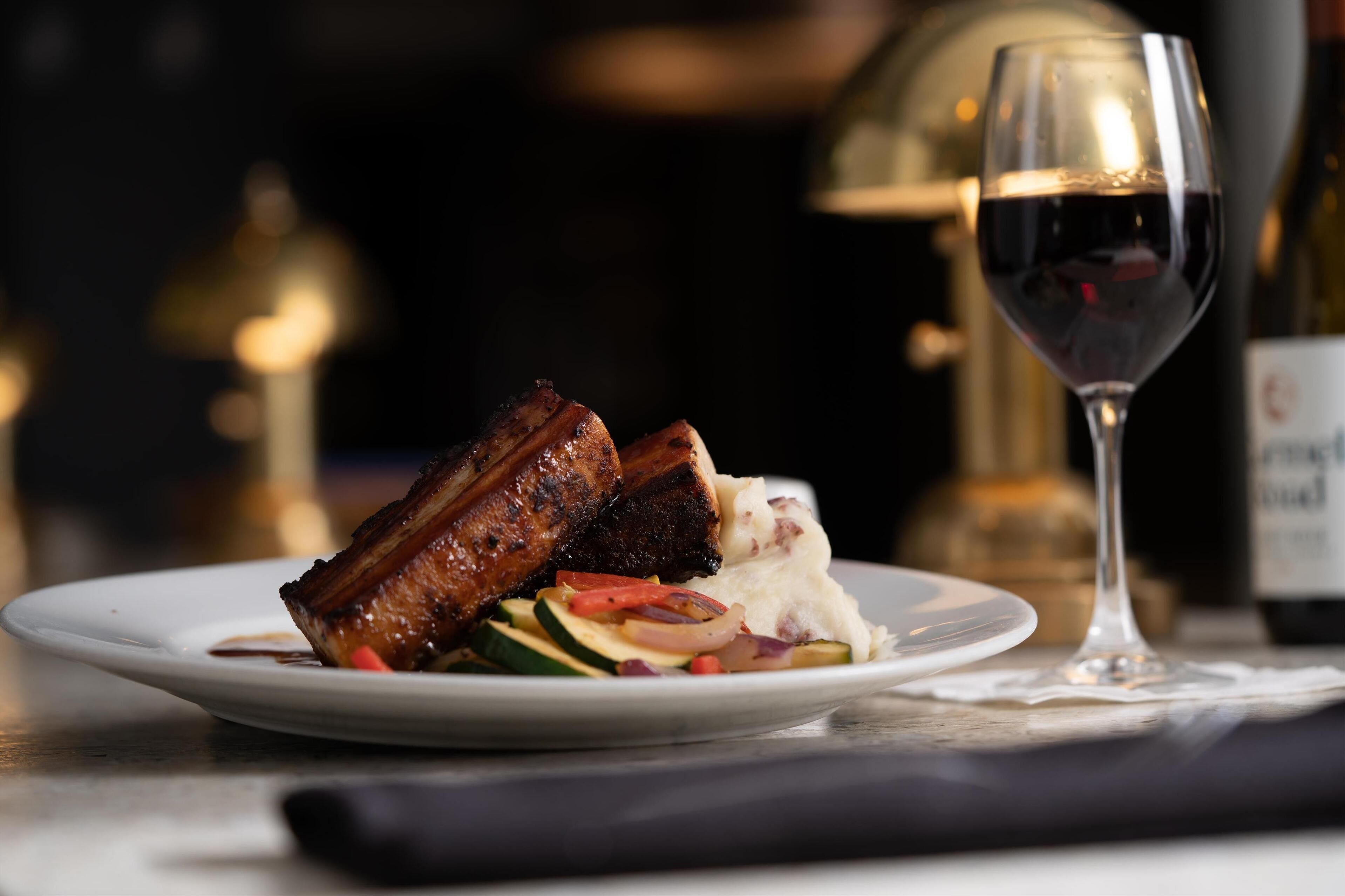 Um prato de comida com carne grelhada e legumes, um copo de vinho tinto e uma garrafa ao fundo.