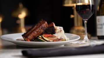 Um prato de comida com carne grelhada e legumes, um copo de vinho tinto e uma garrafa ao fundo.