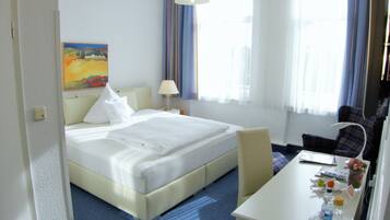 Quarto de hotel com cama, escrivaninha, cadeira, abajur, um quadro na parede e janela com cortinas.