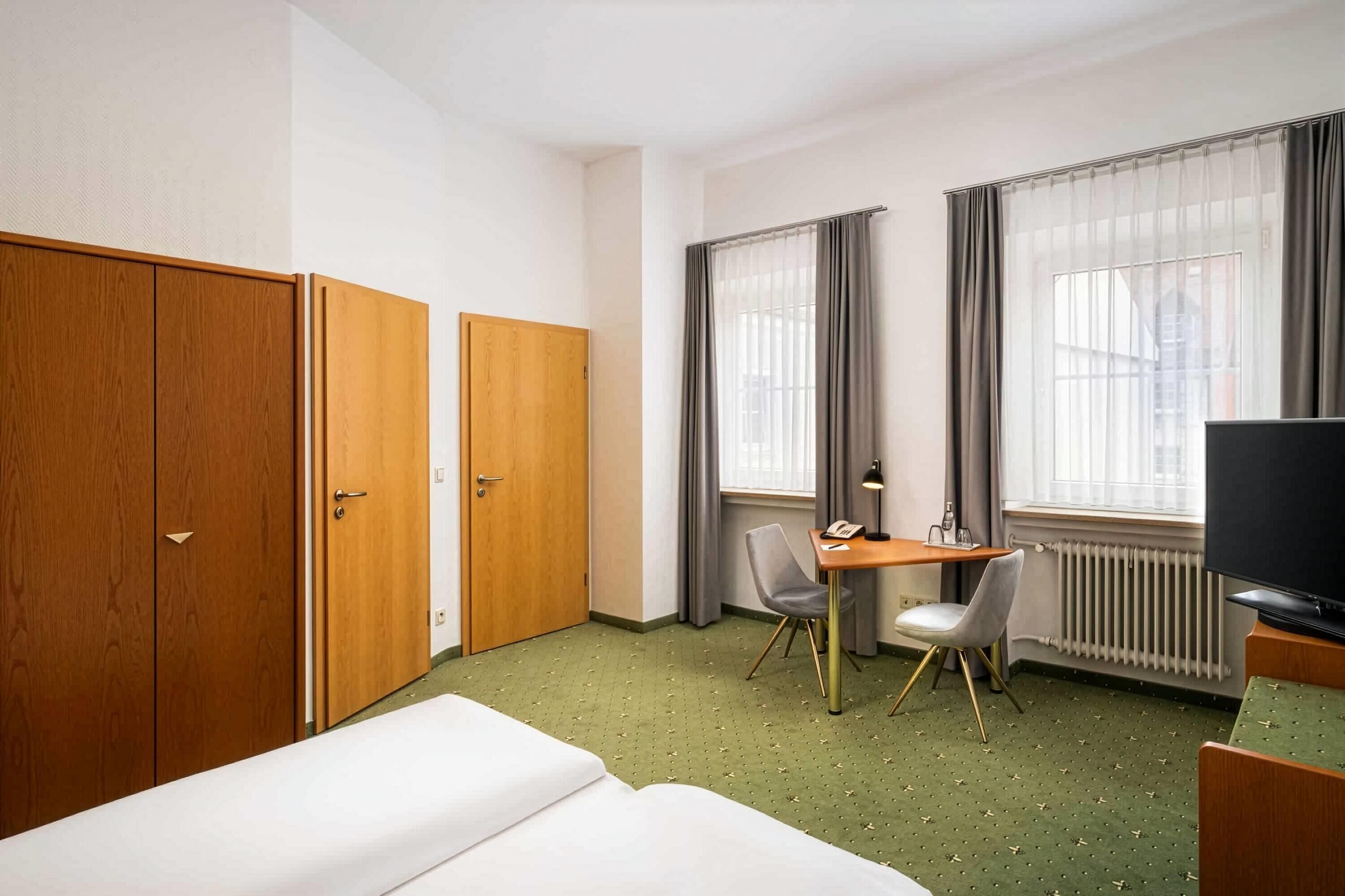 Quarto de hotel com cama, escrivaninha, cadeira, televisão e janela com cortinas.