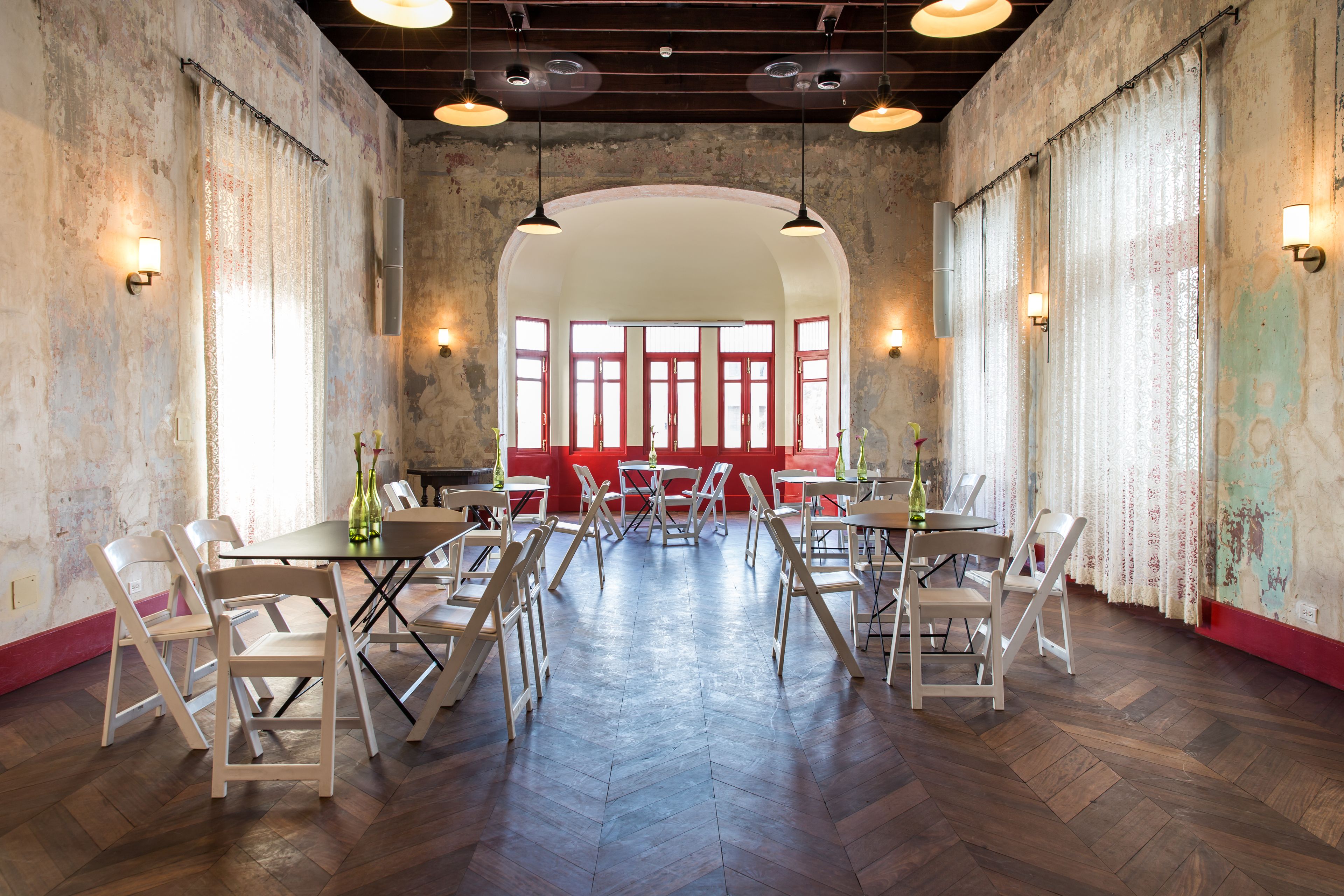 Ampla sala de jantar com piso de madeira em padrão herringbone, cadeiras brancas e mesas dispostas ao redor de um sofá vermelho central.