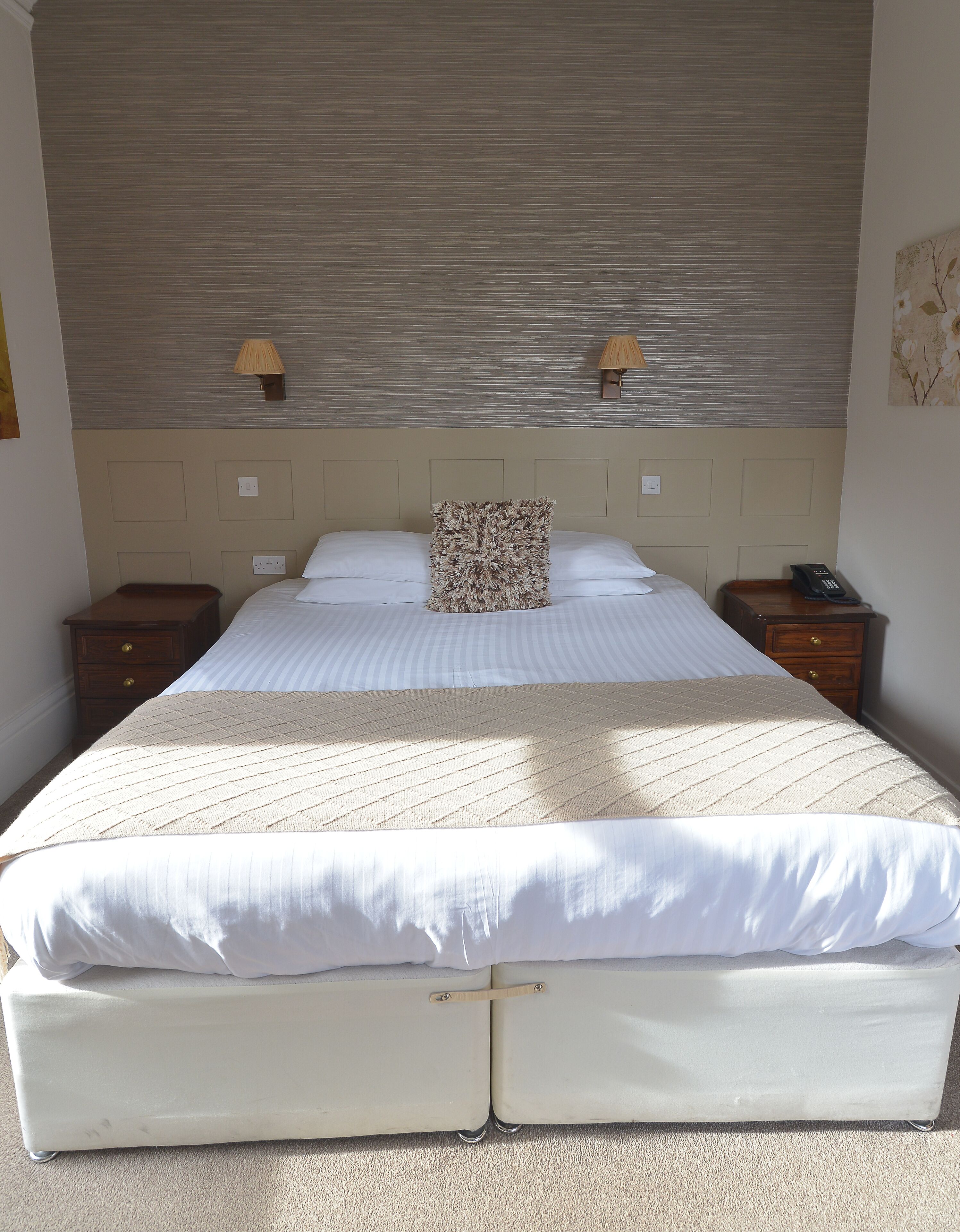 Cama bem arrumada com travesseiro estampado, duas mesas de cabeceira com abajures e luminária fixada na parede.