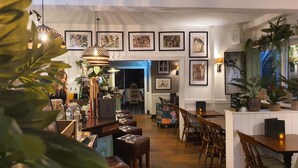 Interior de restaurante com mesas e cadeiras de madeira, luminárias pendentes e quadros nas paredes.