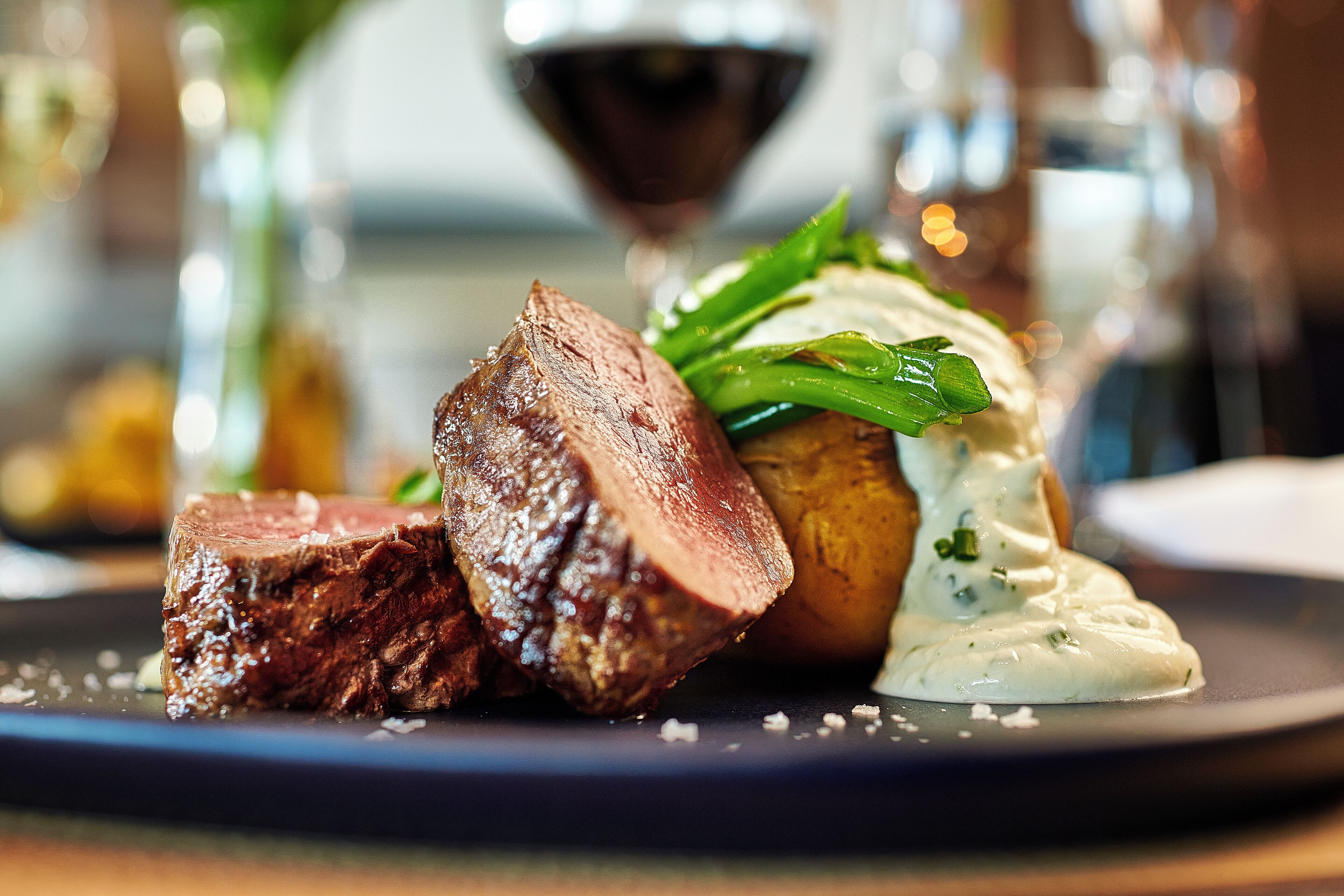 Um prato com bife, batata e molho cremoso, com um copo de vinho tinto ao fundo.