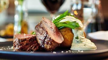Um prato com bife, batata e molho cremoso, com um copo de vinho tinto ao fundo.