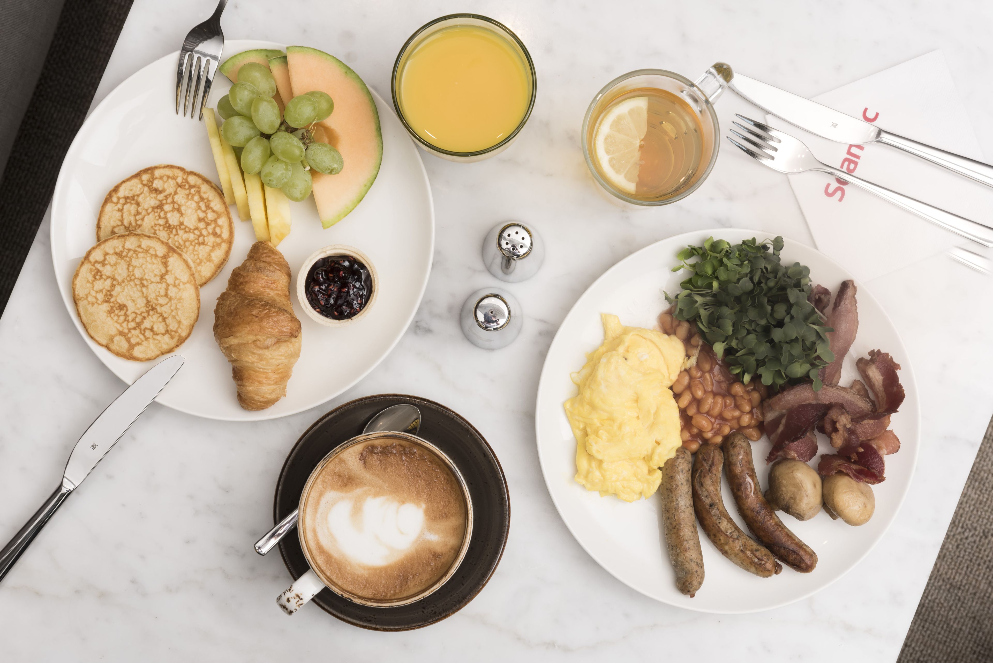Mesa de café da manhã com panquecas, croissants, café e diversos tipos de frutas e vegetais.