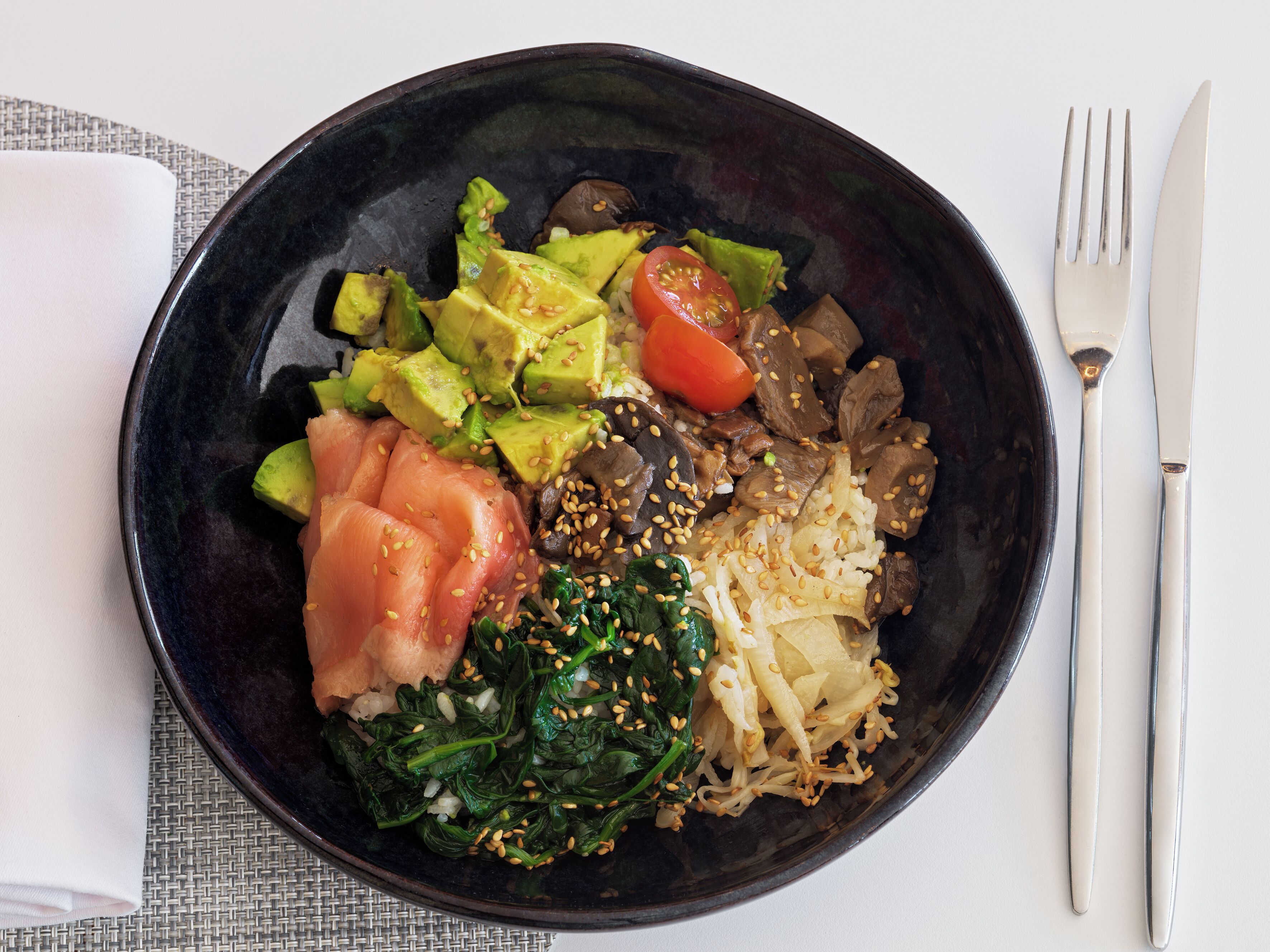 Uma tigela preta com diversos alimentos como abacate, salmão e vegetais, com um garfo e uma faca ao lado.