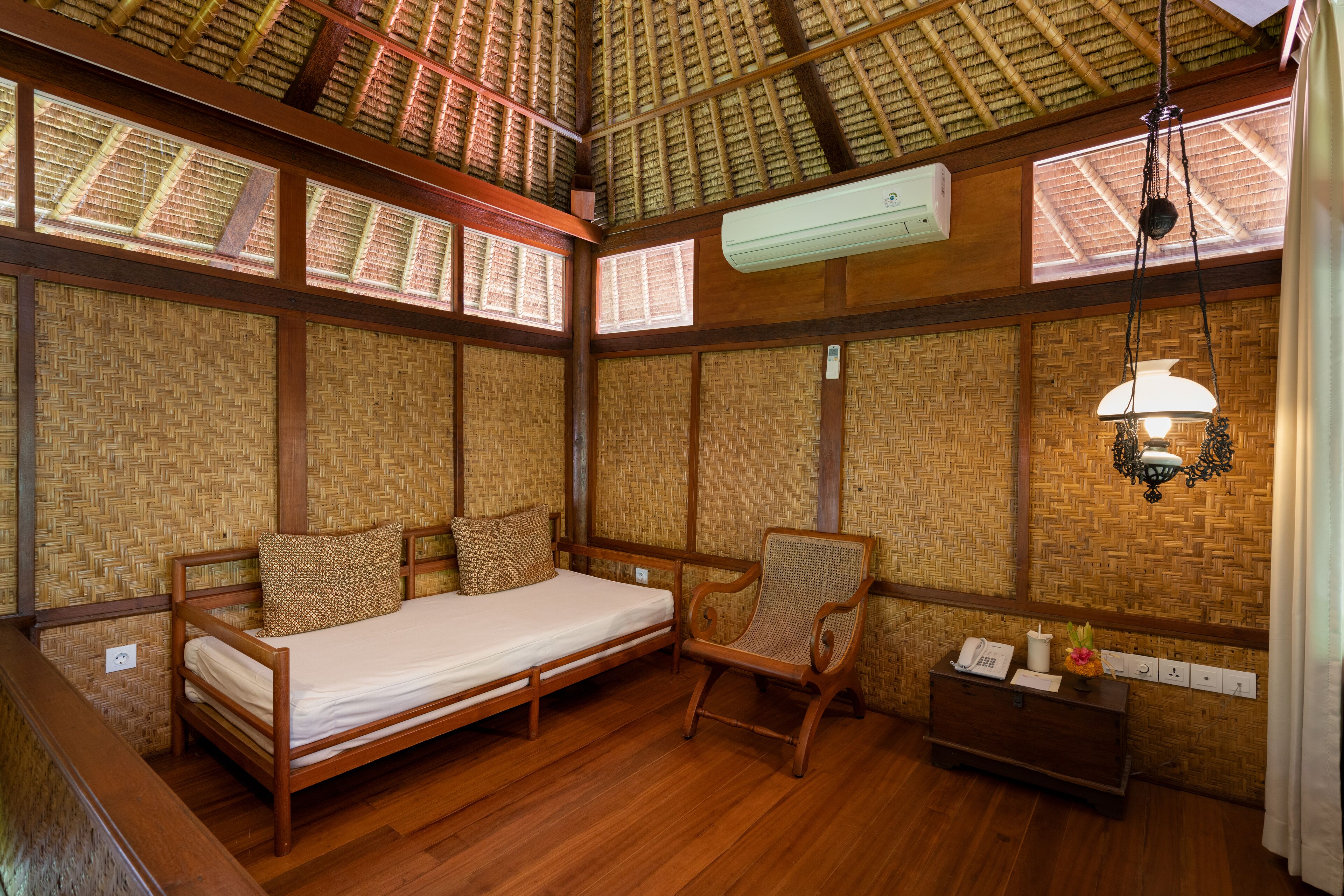 Quarto com cama de madeira, uma cadeira, uma mesinha com telefone e um vaso, e luminária pendente.