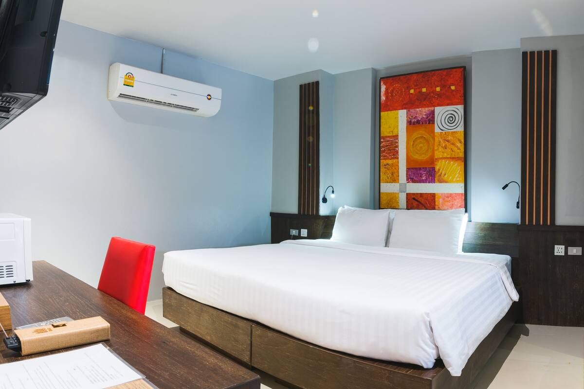 Quarto de hotel com cama, escrivaninha, cadeira, ar condicionado e parede com obra de arte colorida.