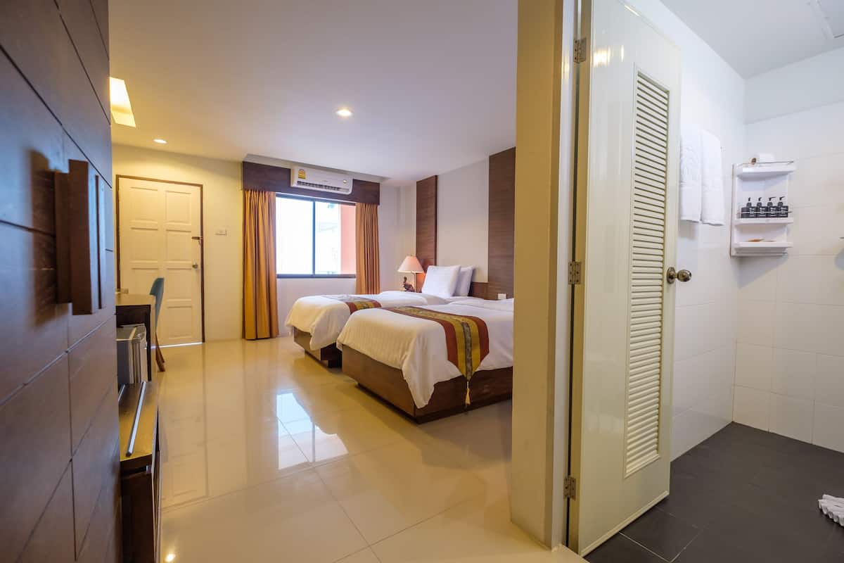 Quarto de hotel com cama, escrivaninha, cadeira, janela com cortinas, banheiro com chuveiro e prateleira com artigos de higiene pessoal.