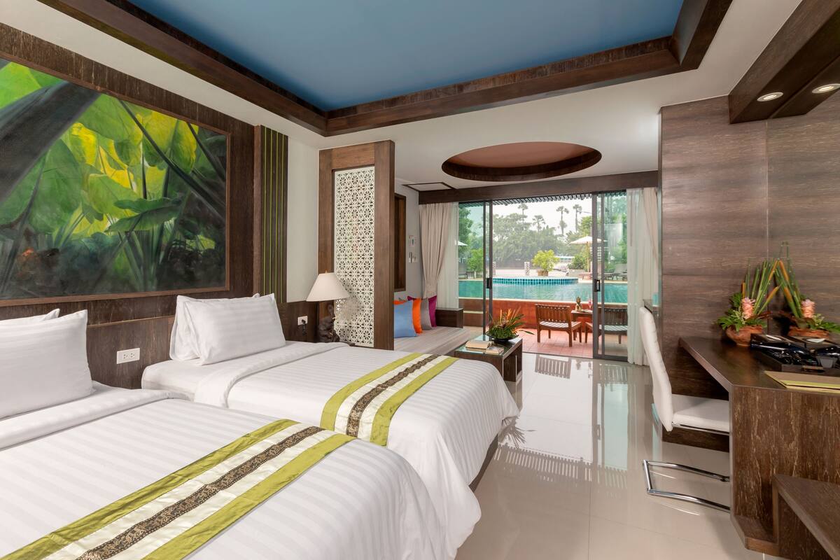 Quarto de hotel com duas camas grandes, uma escrivaninha, uma cadeira, uma mesa de jantar e vista para a piscina e vegetação.