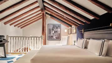Quarto com uma cama grande, corrimão branco, teto com vigas de madeira aparentes e uma obra de arte abstrata emoldurada na parede.