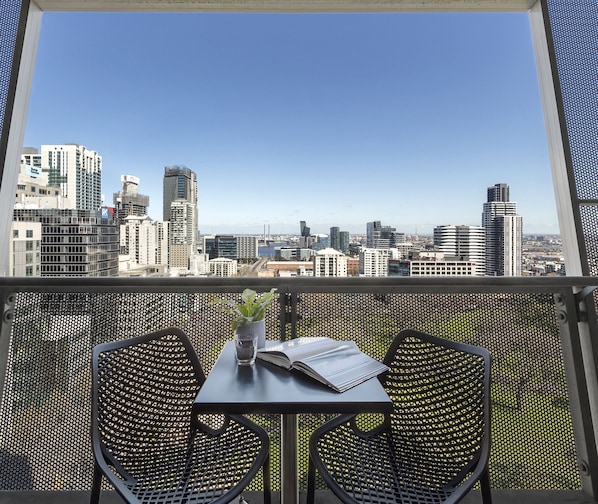 Varanda com duas cadeiras e uma mesa, com vista para uma paisagem urbana com diversos edifícios.