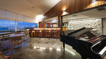 Uma área de bar moderna com um piano de cauda, painéis de madeira e vista para a paisagem urbana.