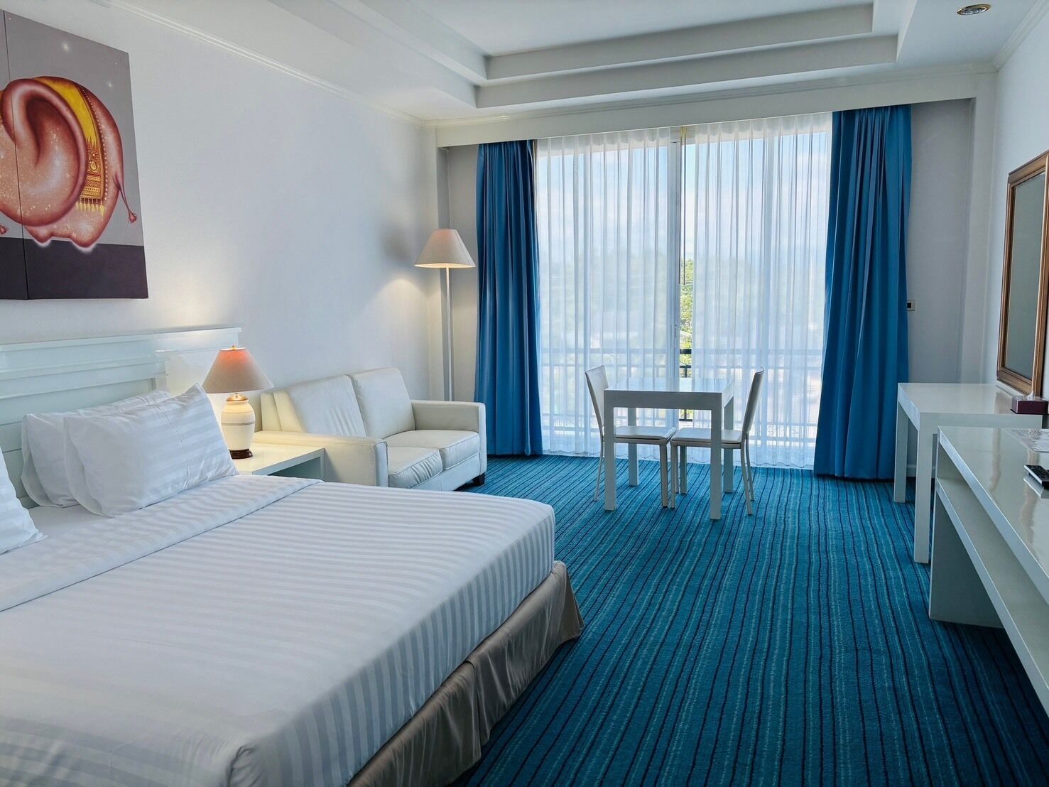 Quarto de hotel com uma cama grande, uma mesa de trabalho, uma cadeira e vista através de cortinas leves.