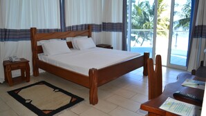 Quarto com cama de madeira, mesa de cabeceira, escrivaninha com cadeira e vista para palmeiras pela janela.