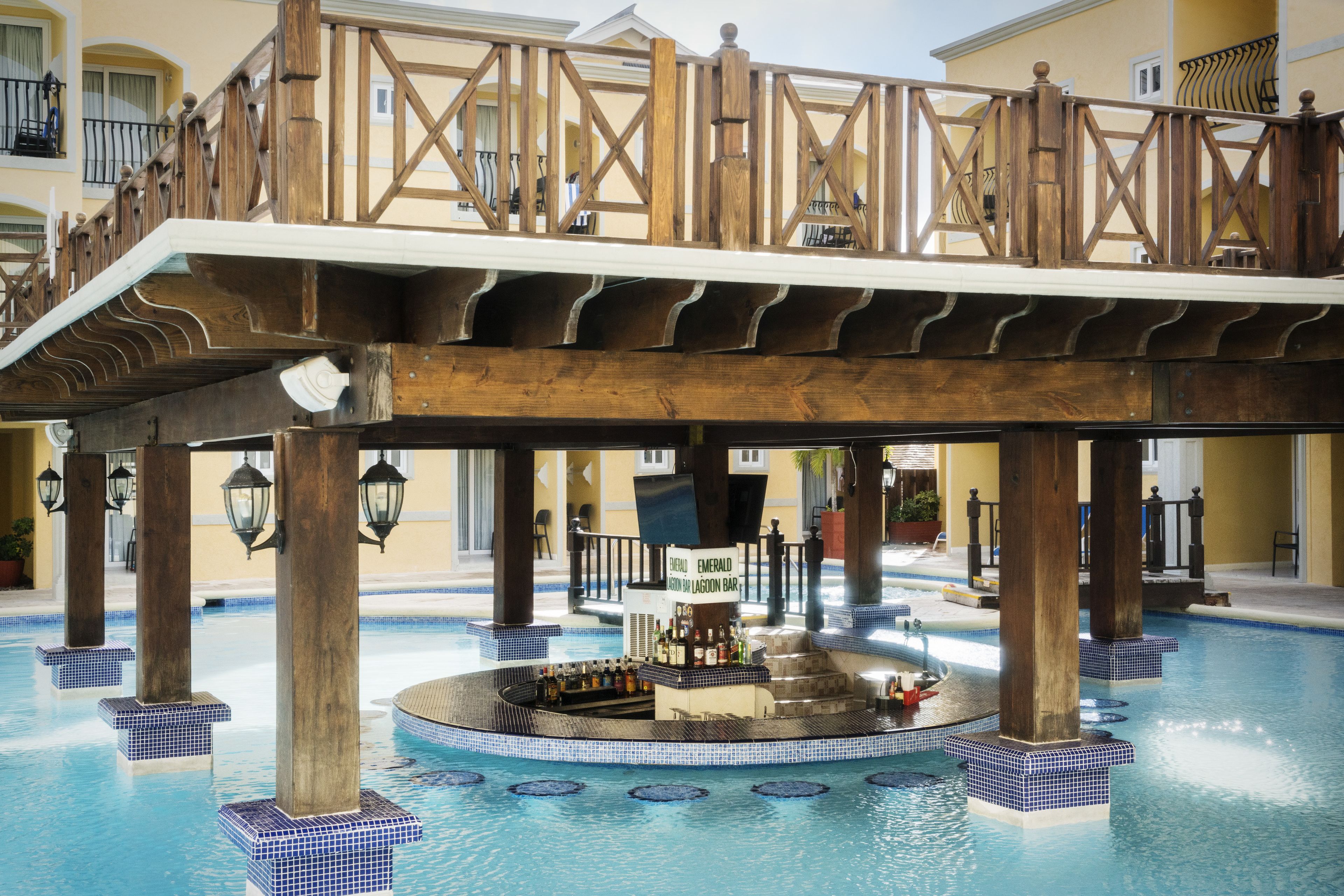 Uma área de piscina com bar sob uma ponte de madeira, sustentada por colunas, e piso de azulejos ao redor da piscina.