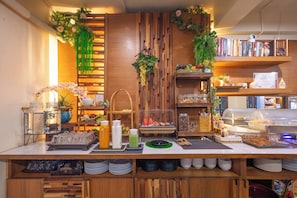 Disposição de buffet com diversos alimentos, um espaço para café e uma seleção de livros em estantes.