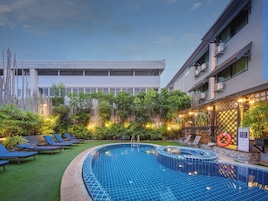 Piscina com borda azulejada, rodeada por espreguiçadeiras e vegetação, com um edifício ao fundo.