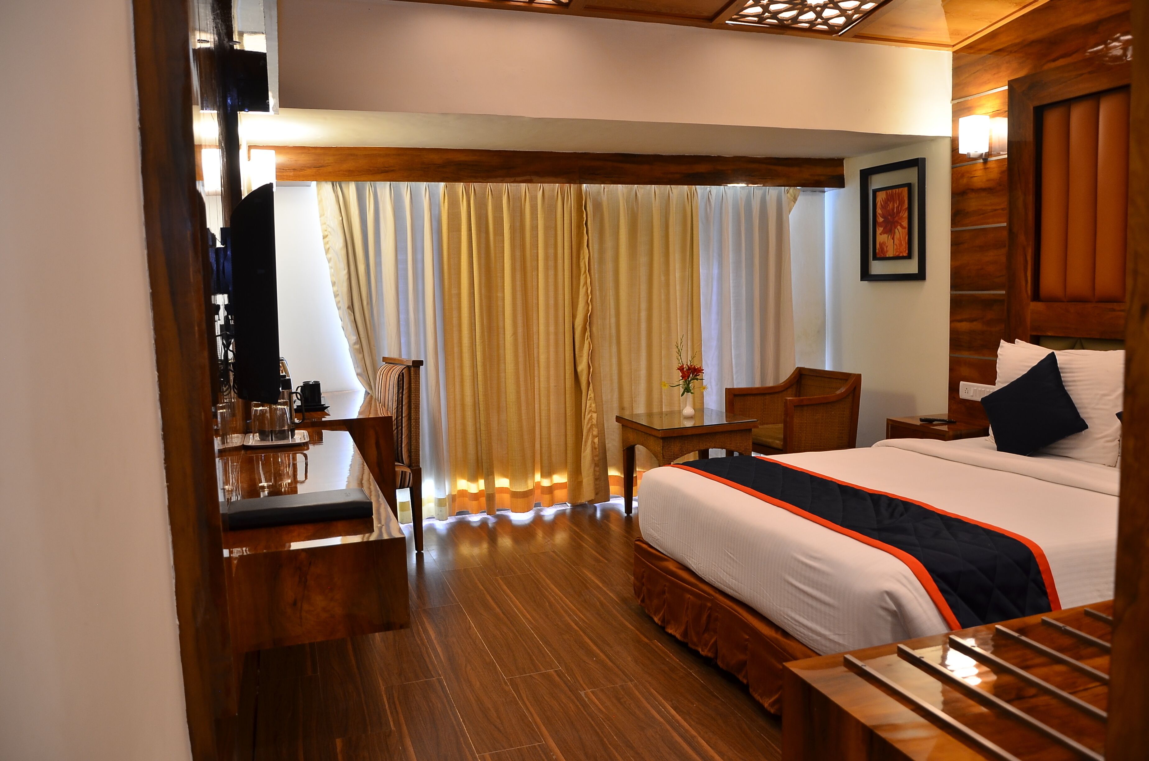 Quarto de hotel com piso de madeira, cama com colcha branca e azul marinho, uma mesinha com um vaso, uma cadeira e um criado-mudo de madeira.