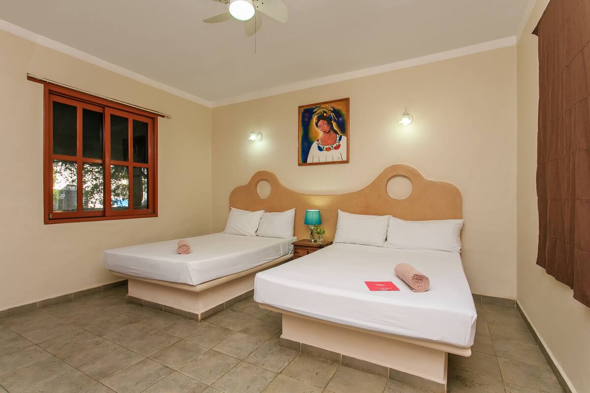 Quarto de hotel com duas camas de solteiro, ventoinha no teto, janela com persianas, quadro na parede e mesa de cabeceira com abajur.