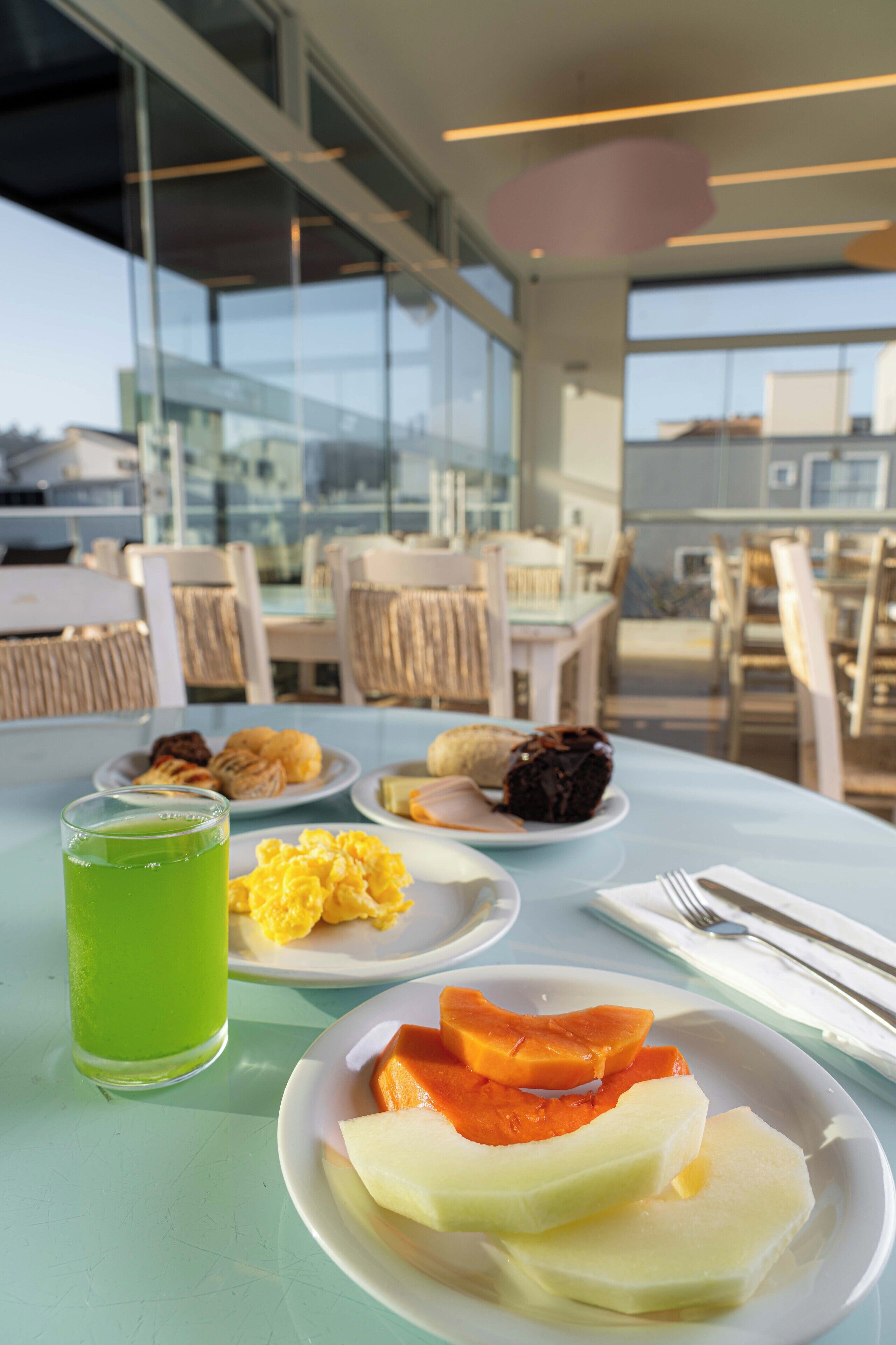 Mesa de jantar com pratos de comida, um copo de bebida verde e vista para a cidade através de grandes janelas.