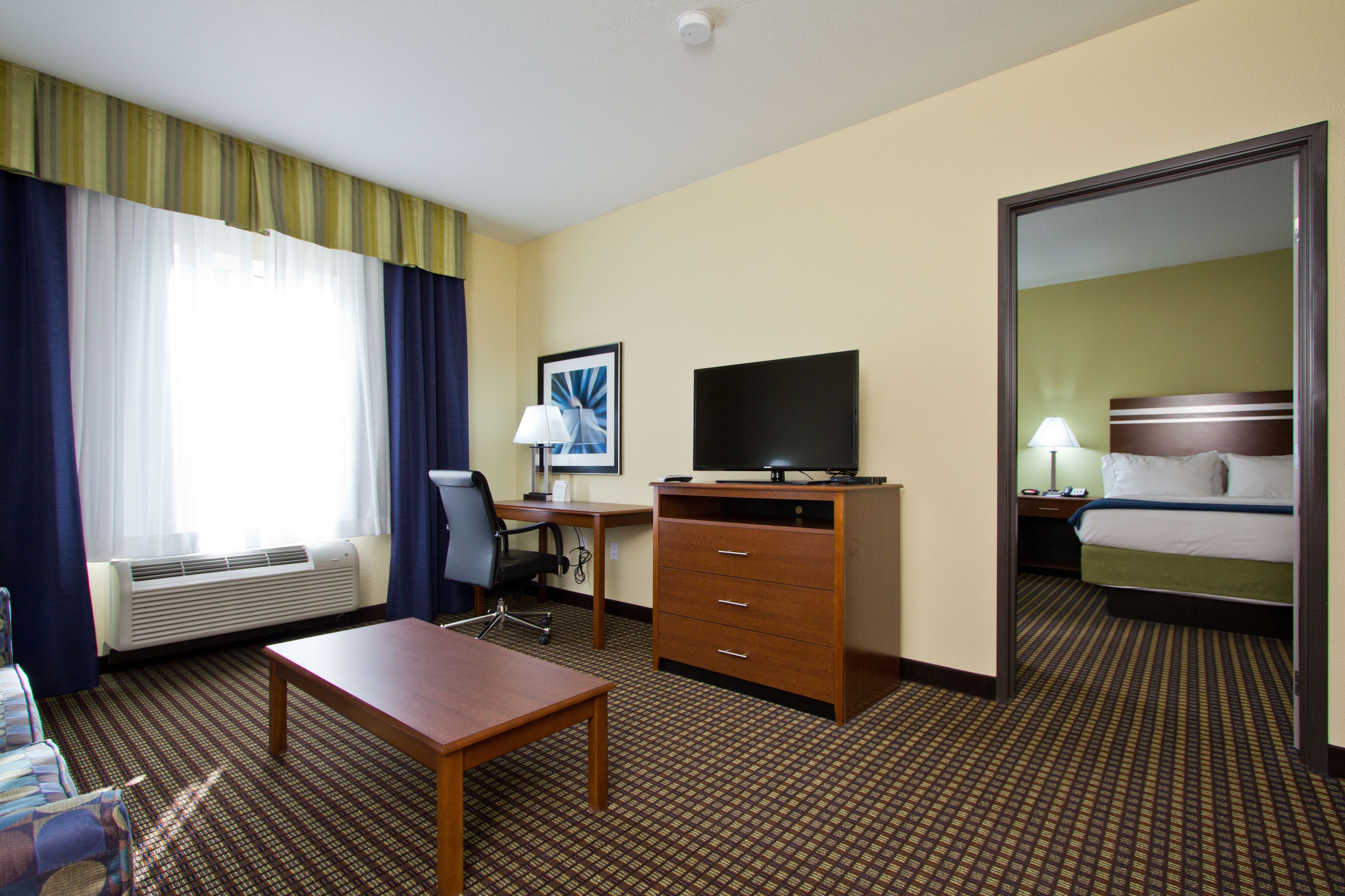 Quarto de hotel com cama, escrivaninha, cadeira, televisão e uma janela grande com cortinas.