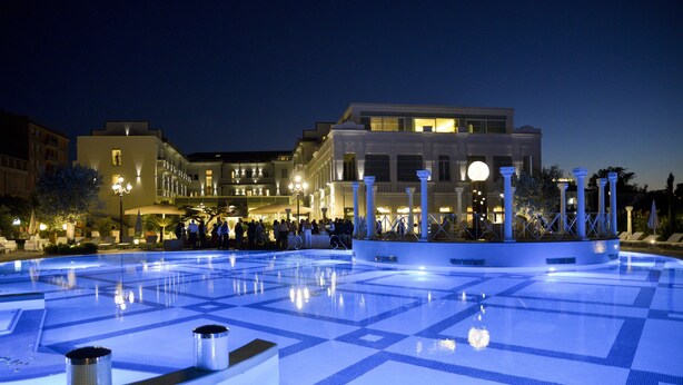 Piscina ao ar livre com padrão geométrico de azulejos, cercada por edifícios com varandas iluminadas e uma fonte no centro.
