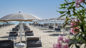 Uma praia com fileiras de espreguiçadeiras e guarda-sóis, alguns com a sinalização 'GRAND HOTEL da VINCI'.