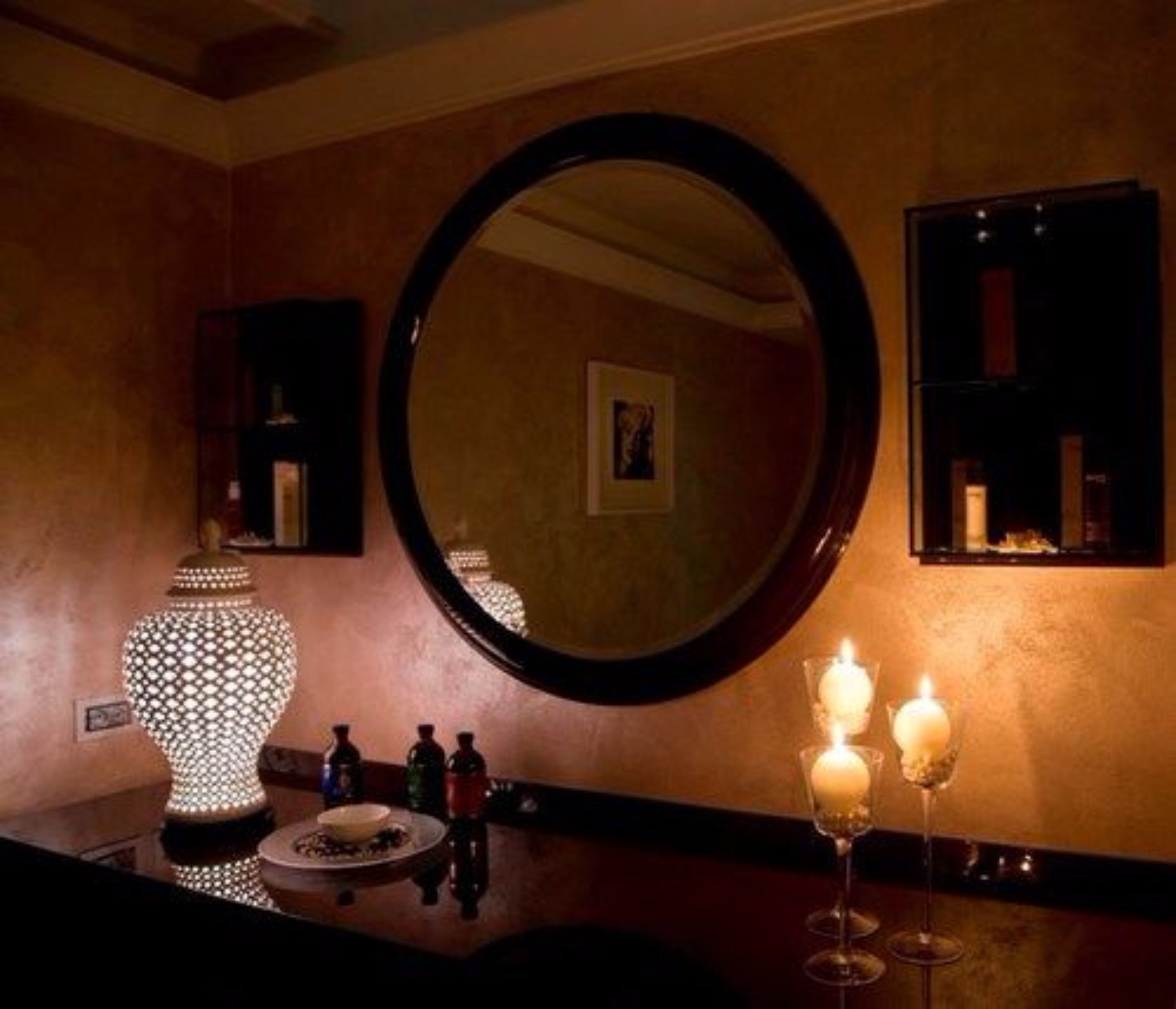 Um quarto com pouca luz, um espelho grande, uma luminária decorativa e três velas acesas sobre uma mesa.