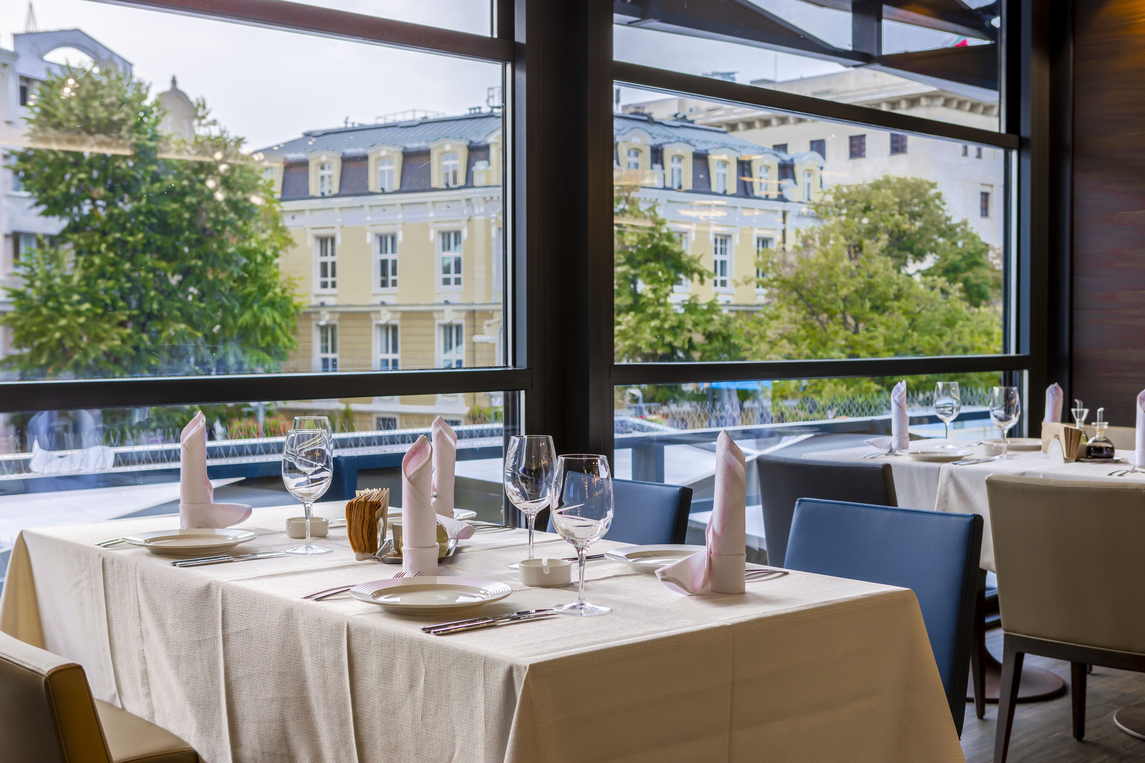 Restaurante com mesas preparadas para refeições, amplas janelas oferecendo vista para edifícios e árvores.