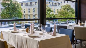 Restaurante com mesas preparadas para refeições, amplas janelas oferecendo vista para edifícios e árvores.