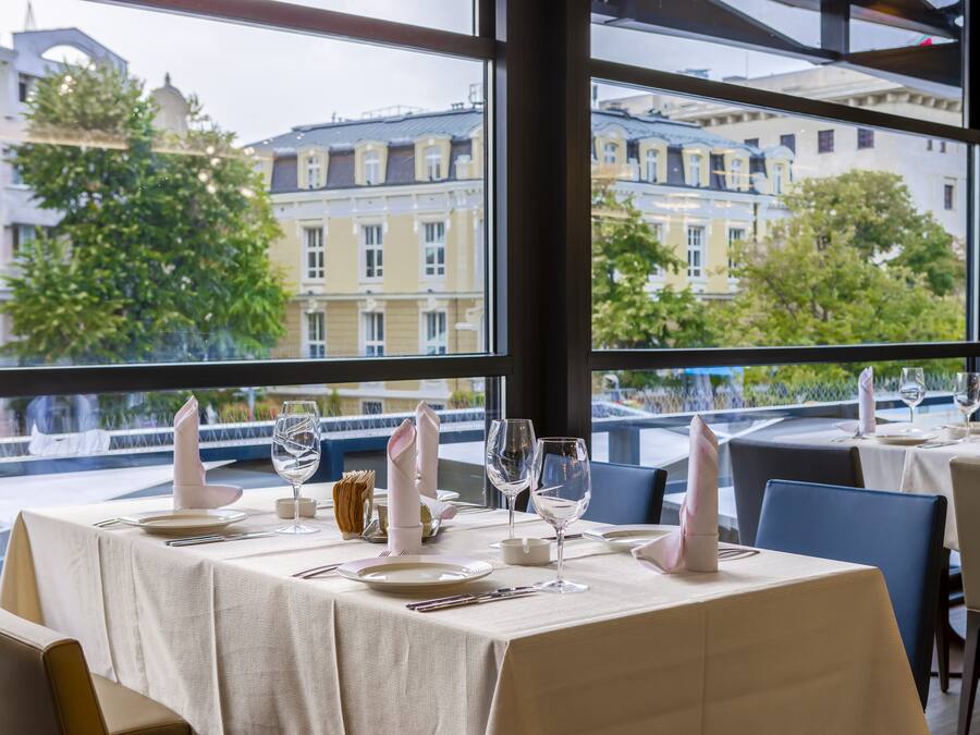 Restaurante com mesas preparadas para refeições, amplas janelas oferecendo vista para edifícios e árvores.