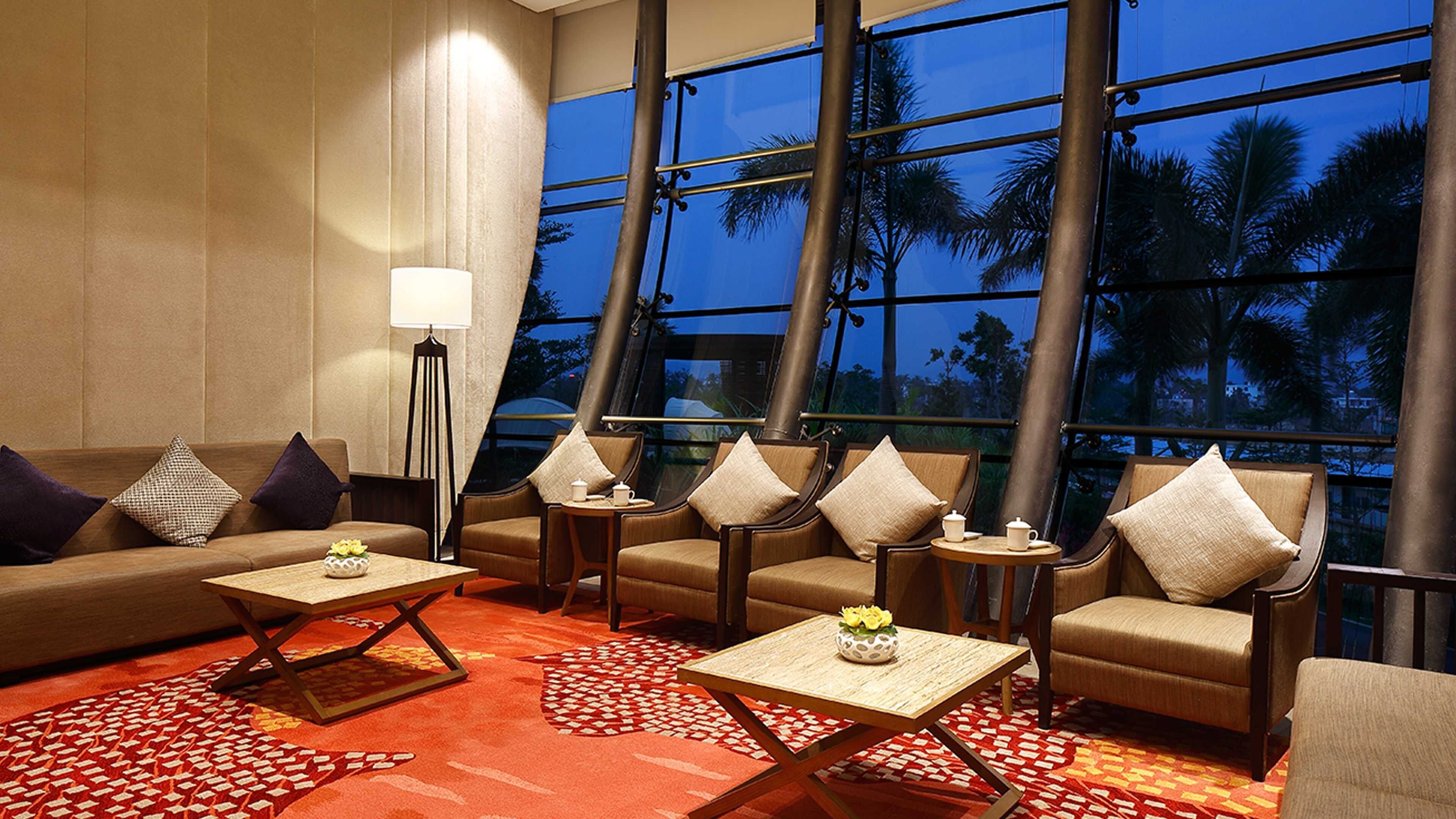 Quarto de hotel com uma grande janela, um sofá, poltronas, uma mesa de centro e um abajur.