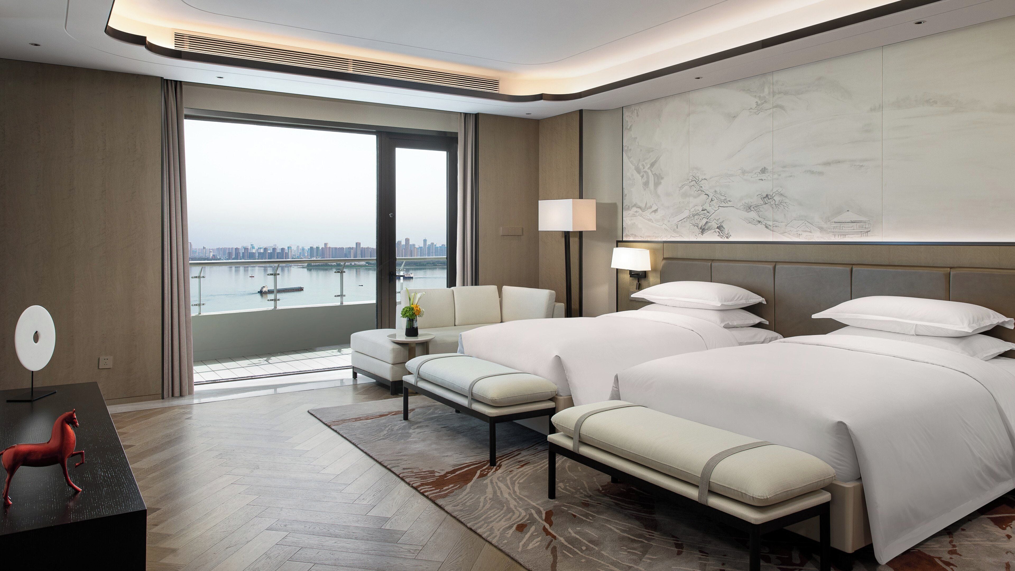 Quarto de hotel com duas camas, um sofá, uma escrivaninha e vista para o horizonte da cidade.