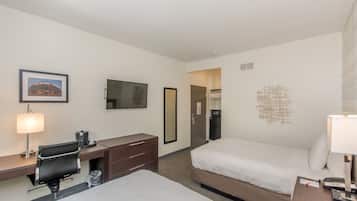 Quarto de hotel com duas camas, uma mesa com cadeira, televisão e banheiro visível através de uma porta aberta.