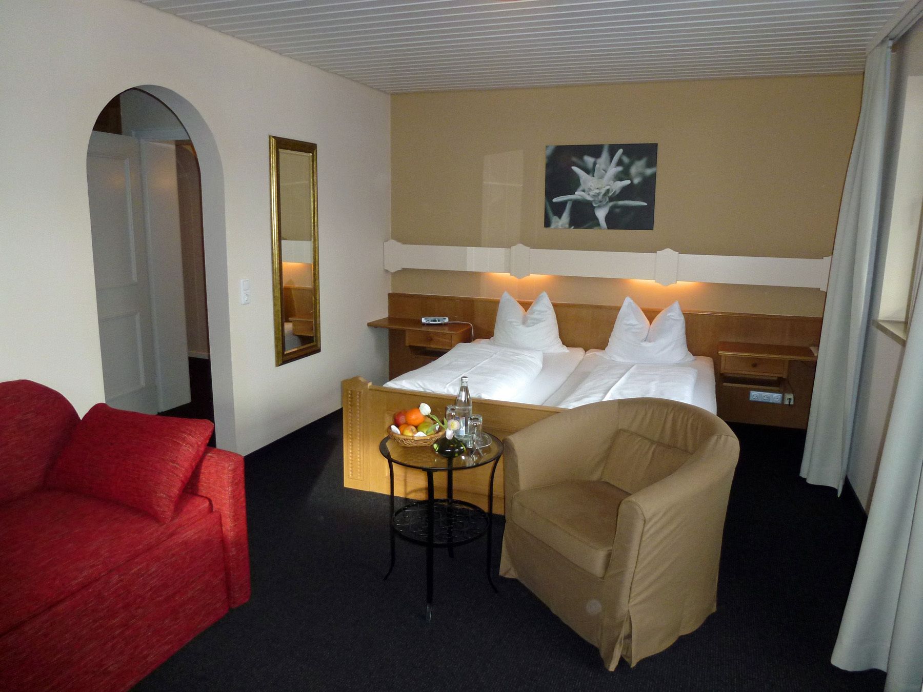 Quarto de hotel com cama, poltrona vermelha, poltrona bege, mesa pequena com cesta de frutas e espelho na parede.
