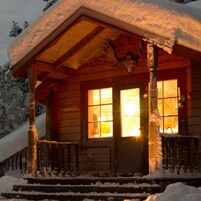 Uma cabana de madeira com telhado coberto de neve e uma janela iluminada, rodeada por neve.