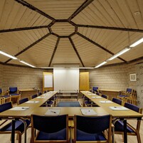 Sala de conferências com paredes e teto de madeira, equipada com mesas, cadeiras, tela de projetor e quadro branco.