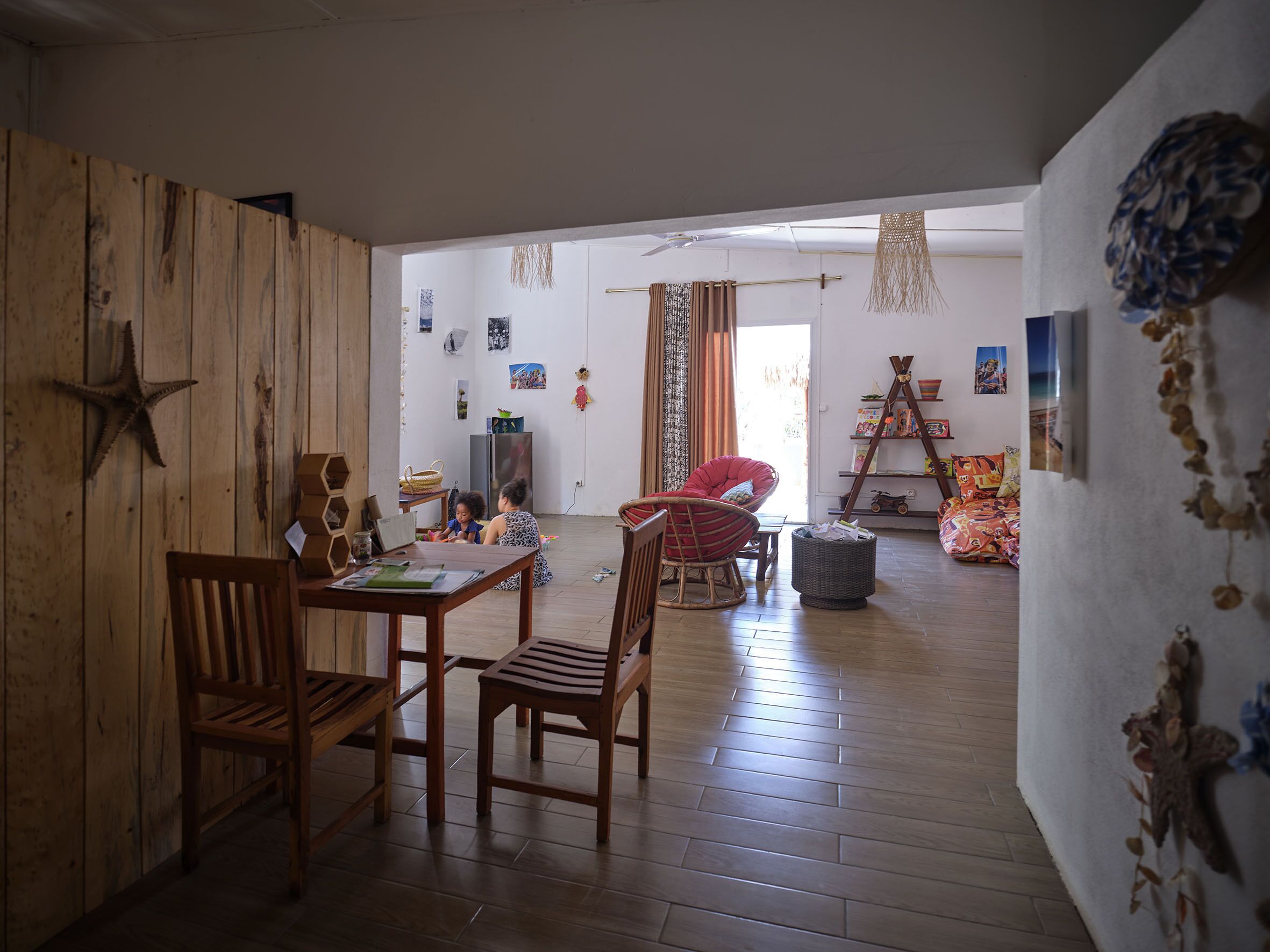 Uma sala de estar ampla com piso de madeira, uma mesa de jantar com cadeiras, uma poltrona vermelha, uma mesinha com objetos e uma estante de livros com brinquedos.