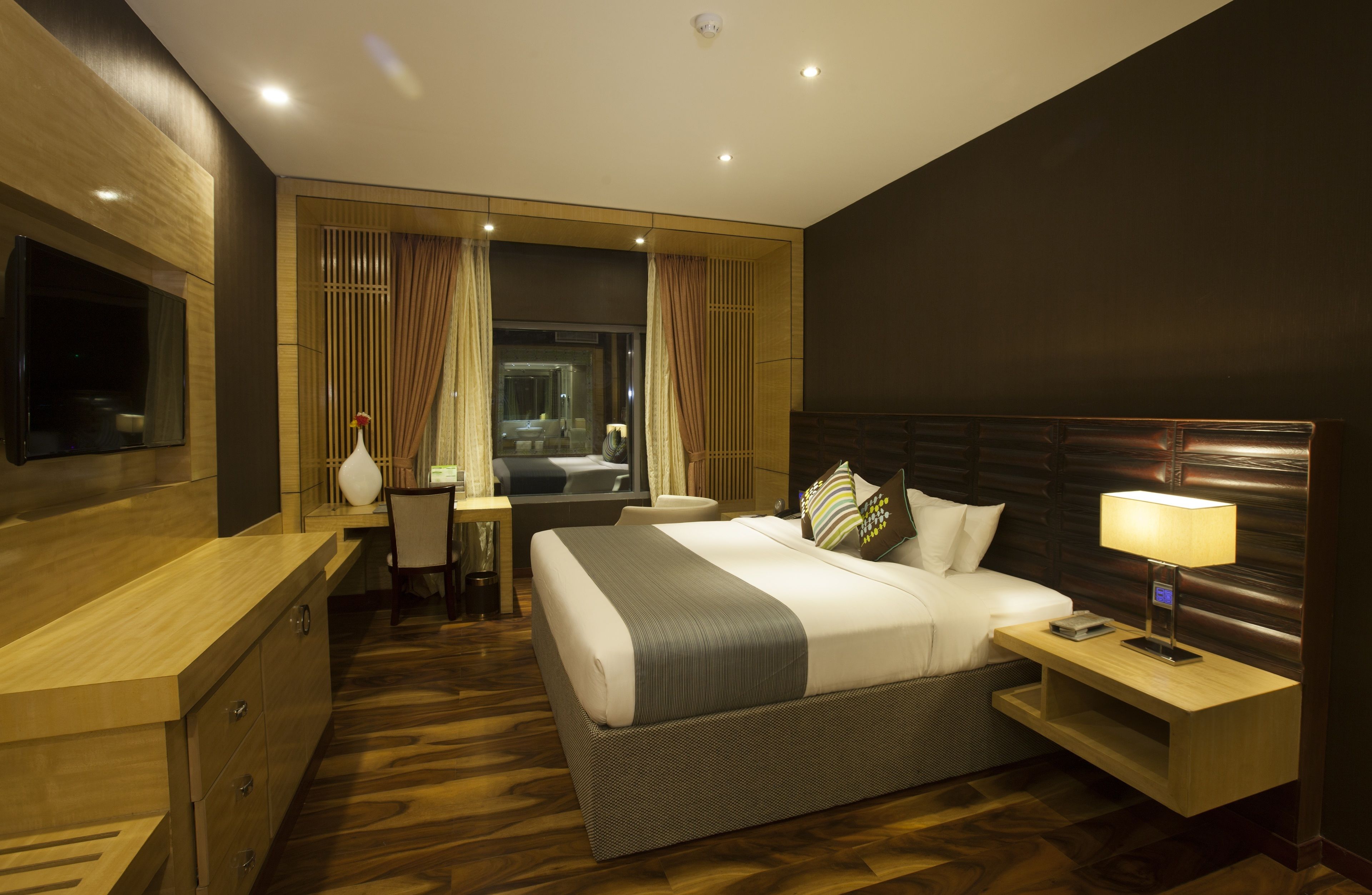Quarto de hotel moderno com uma cama grande, mesa de cabeceira com abajur, escrivaninha com cadeira e televisão na parede.