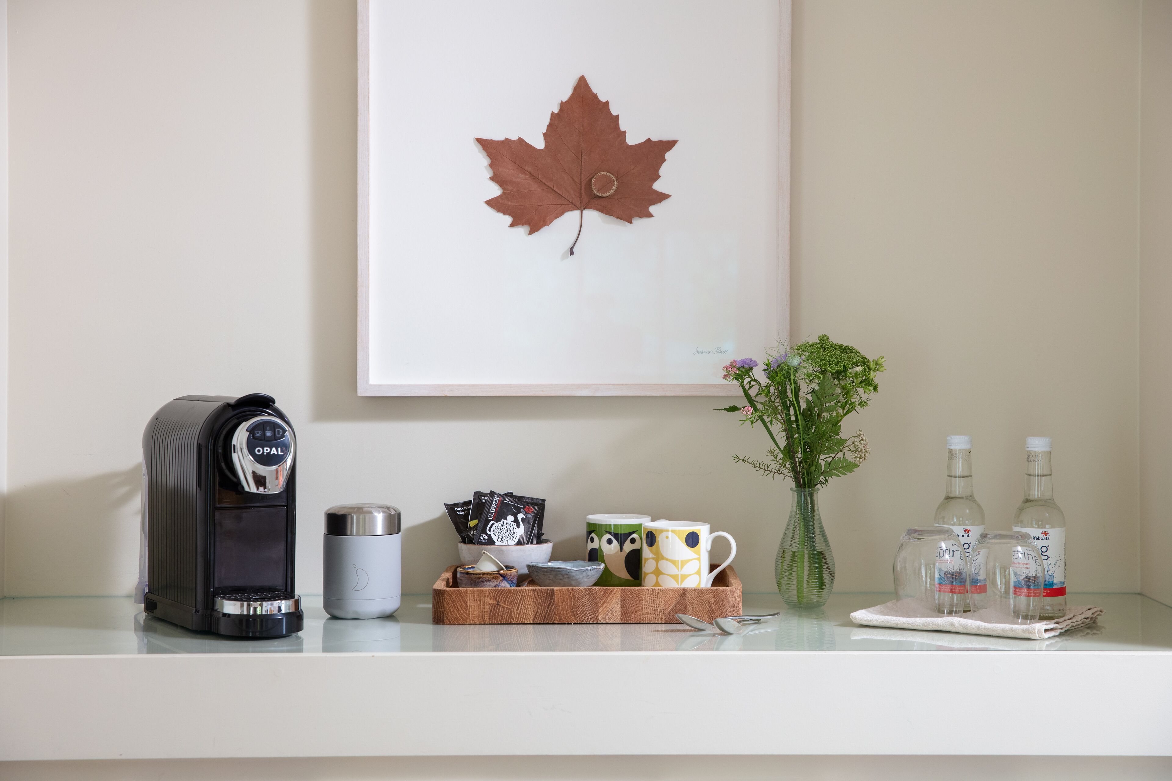 Máquina de café, canecas, bandeja com cápsulas de café, um vaso com flores e garrafas de água sobre uma mesa.