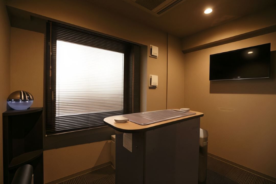 Quarto de hotel com escrivaninha, televisão de tela plana na parede, janela com persianas e uma pequena prateleira com objeto decorativo.