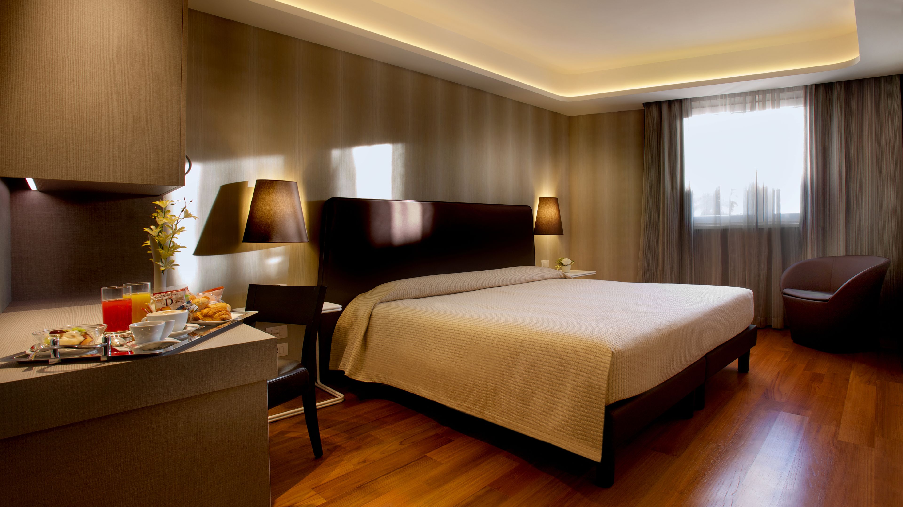 Quarto de hotel com cama, mesas de cabeceira, escrivaninha e objetos de pequeno-almoço, e uma cadeira.