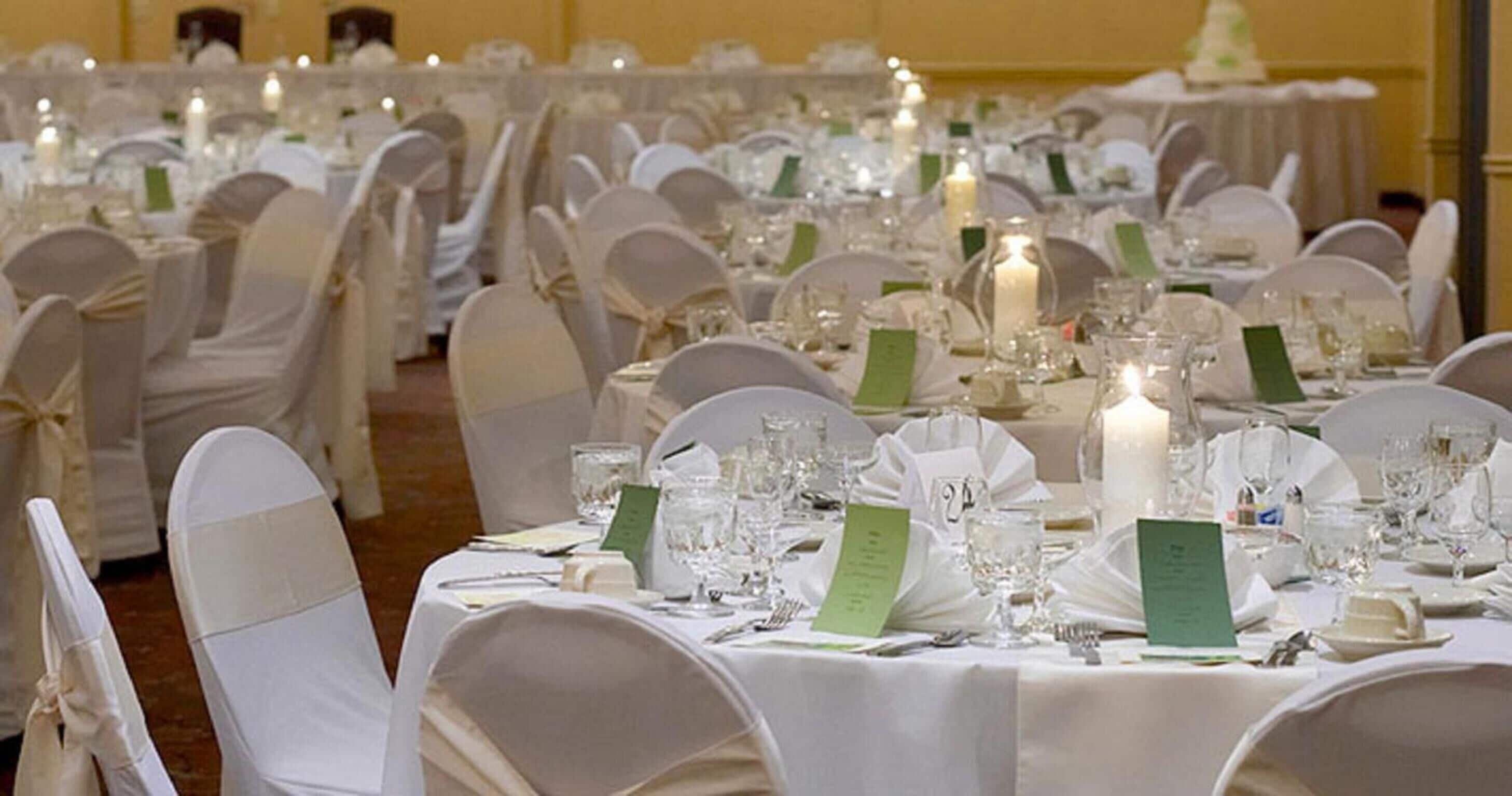 Uma sala de banquetes com mesas redondas preparadas para um evento formal, cada uma com louça posta, velas e cardápios.