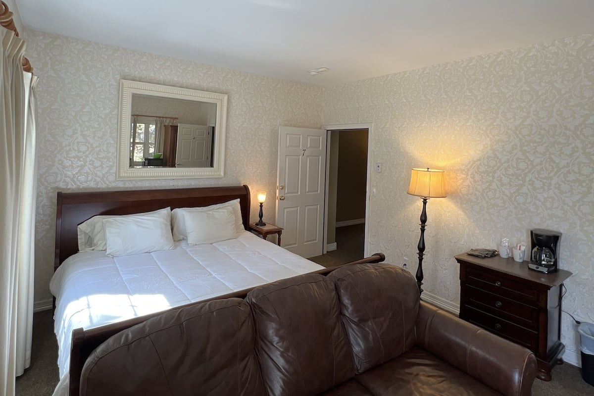 Quarto de hotel com cama grande, sofá de couro marrom, luminária de cabeceira e cômoda com cafeteira.