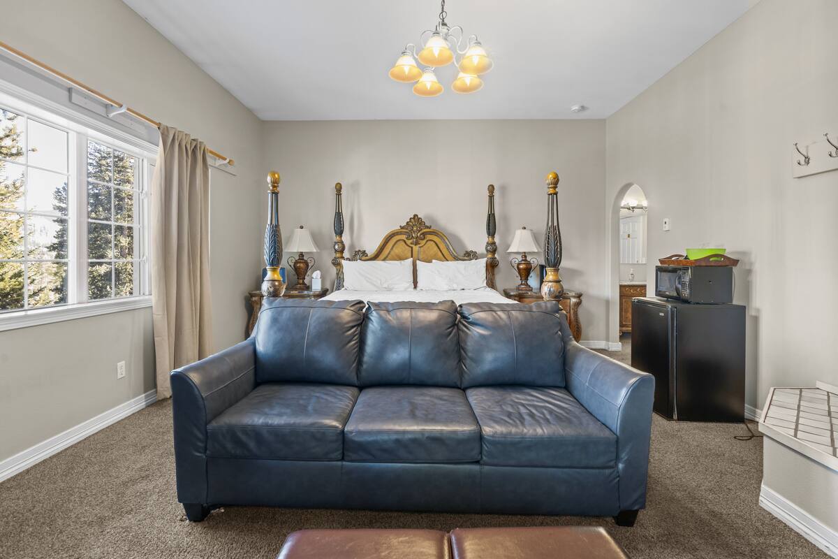 Quarto com um amplo sofá de couro azul, cama com cabeceira ornamentada, lustre e um pequeno frigorífico preto.