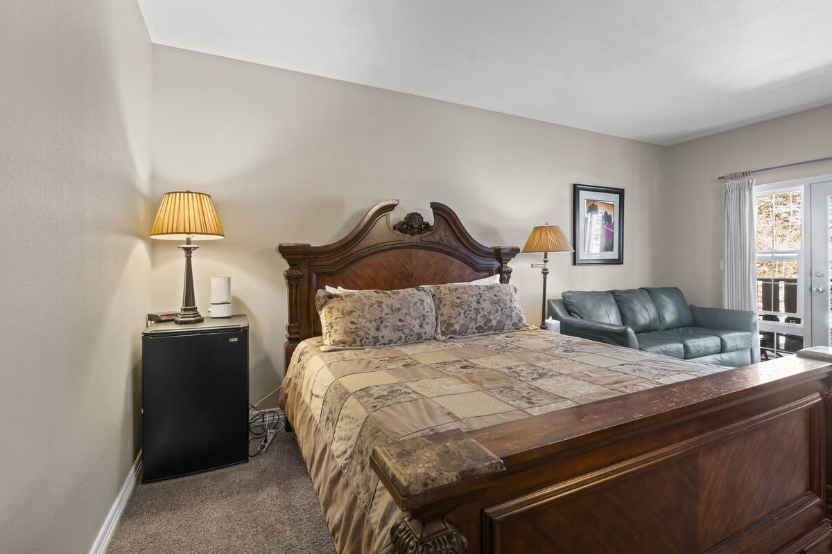 Quarto com cama de madeira, duas luminárias de apoio, um refrigerador preto pequeno, um sofá de couro verde e um quadro pendurado na parede.