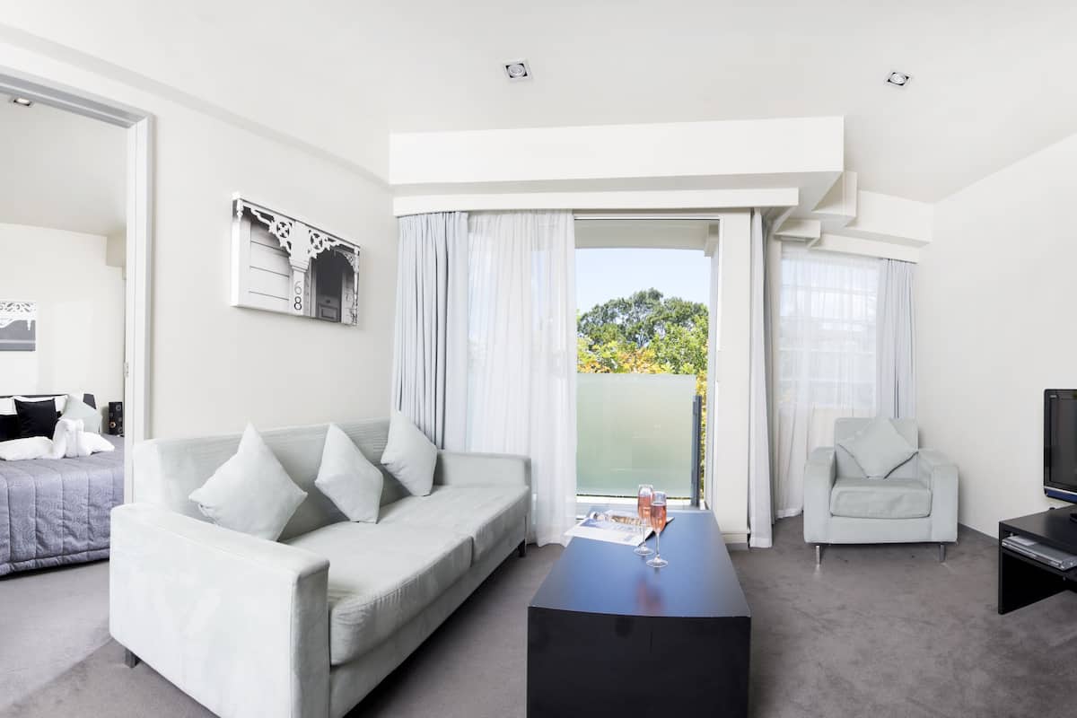 Quarto de hotel moderno com sofá cinzento, poltrona branca, TV de tela plana e varanda com vista para árvores.